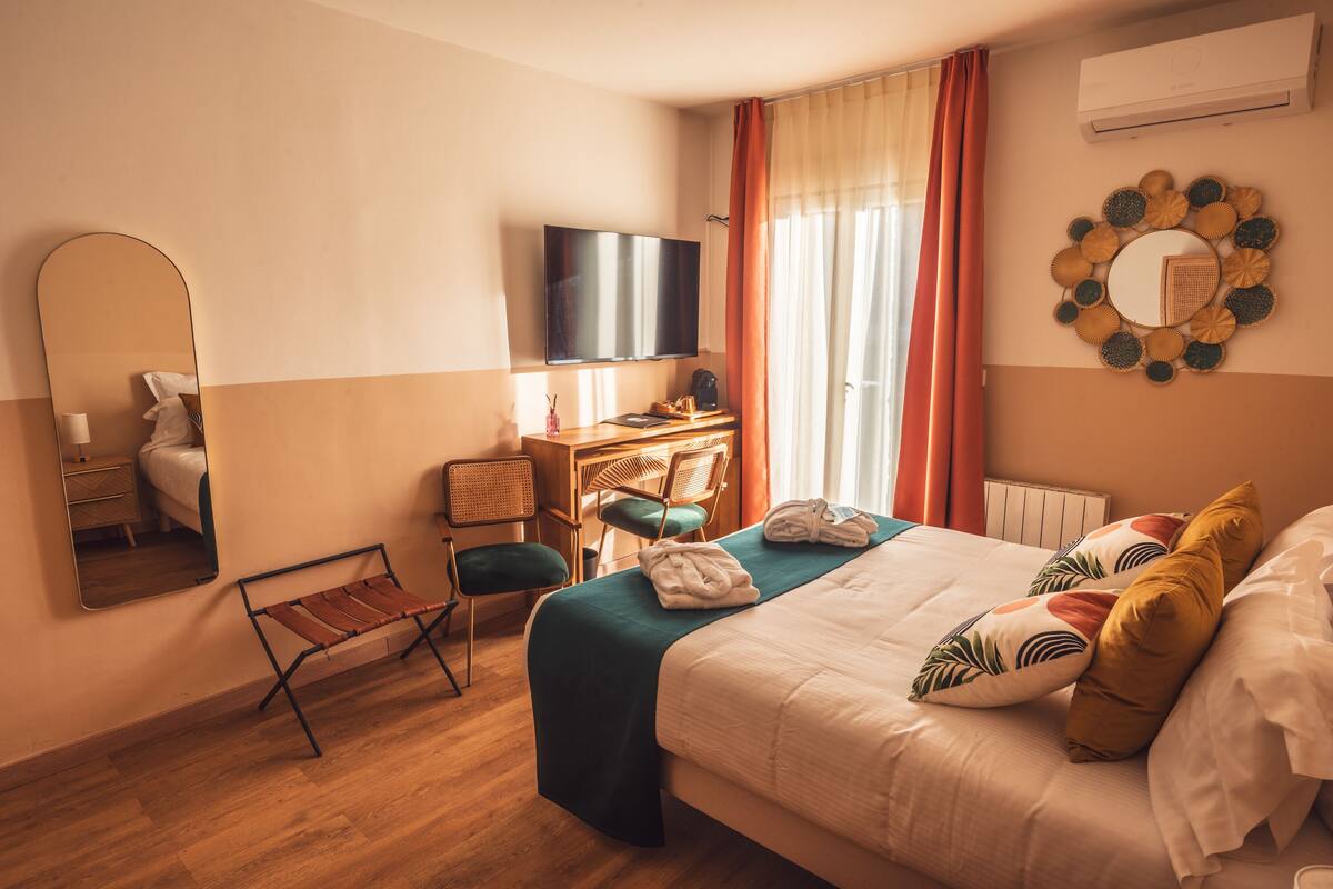 Quarto de hotel com uma cama, escrivaninha, cadeira, televisão e espelho.