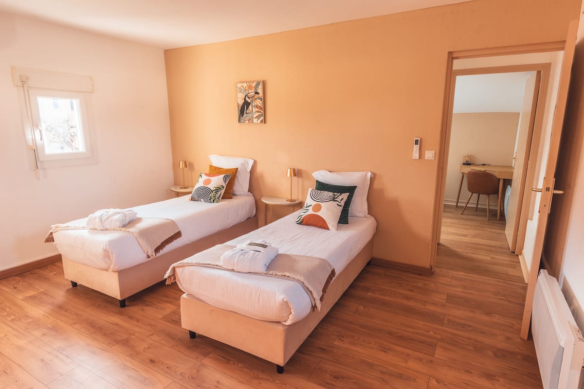 Quarto com duas camas, uma janela, um quadro e uma escrivaninha ao fundo.