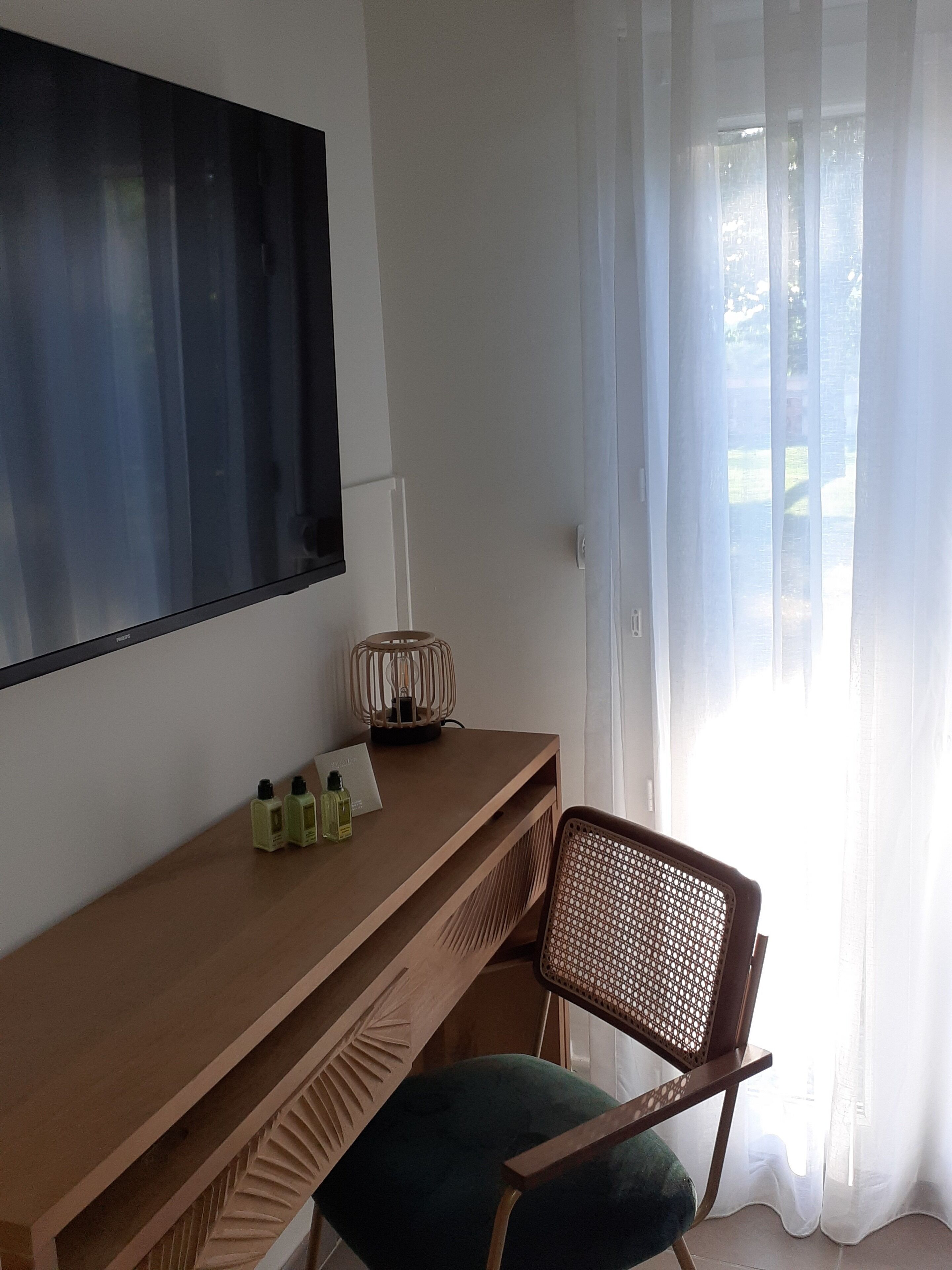 Quarto moderno com escrivaninha de madeira, uma cadeira, TV de tela plana e janela com cortinas leves.