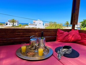 Mesa com bandeja de comida e bebidas, uma garrafa e um par de sapatos numa varanda com vista para a costa.