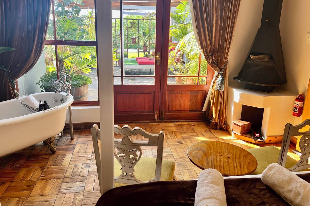 Quarto com piso de madeira, banheira, lareira e vista para um jardim através de uma porta de vidro.
