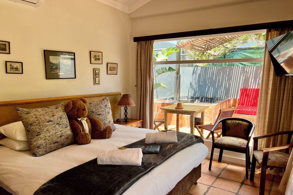 Quarto de hotel com cama, um urso de pelúcia, abajur, televisão e vista para área externa com mesa e cadeiras.