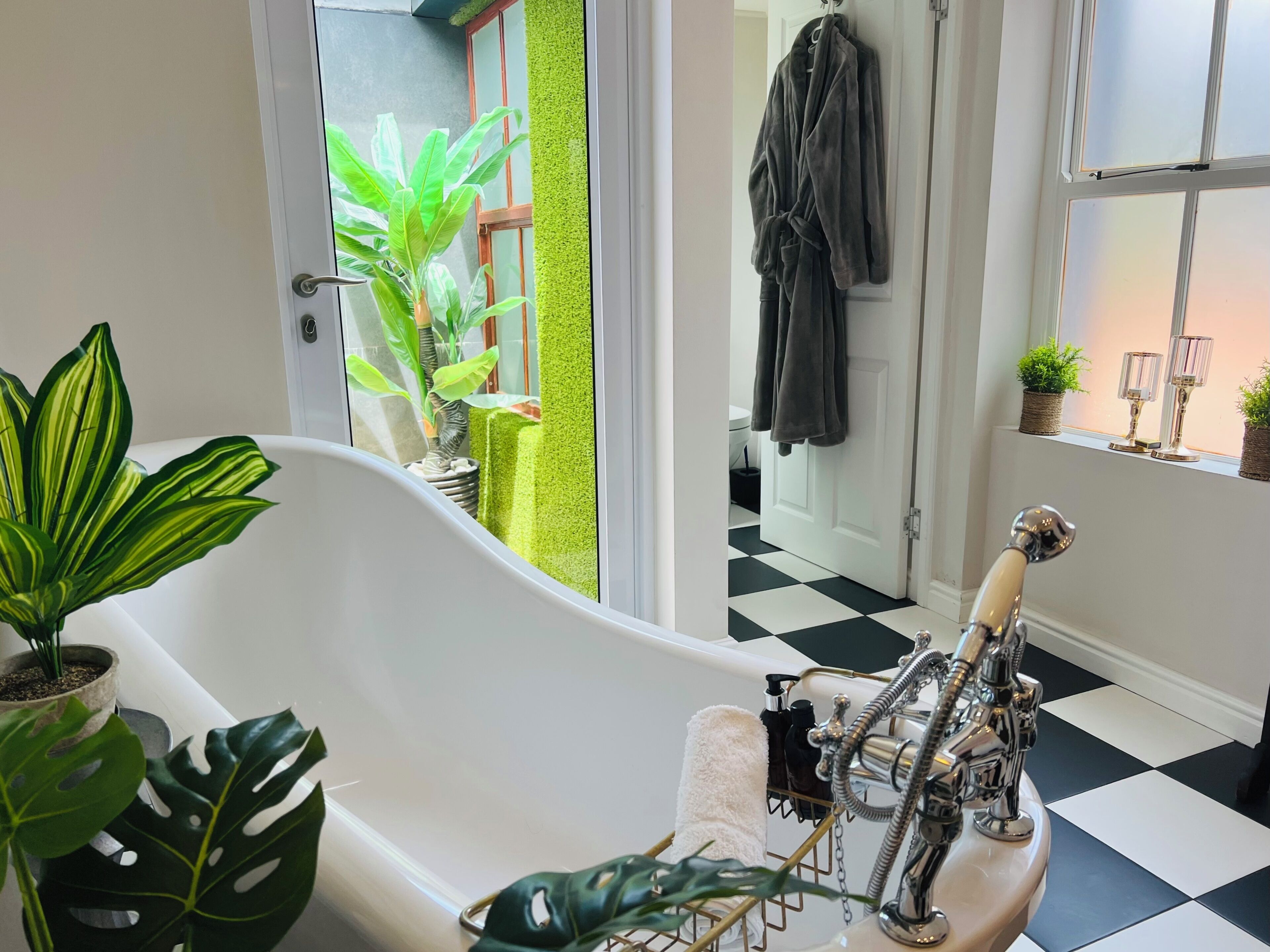 Casa de banho com banheira branca, uma planta verde, um vaso de plantas, um porta-toalhas e um chão xadrez.