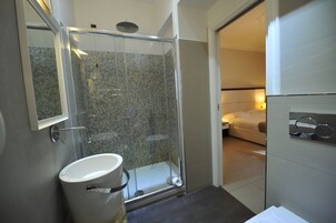Um banheiro moderno com chuveiro em vidro, pia branca e quarto visível, com cama e mesa de cabeceira.