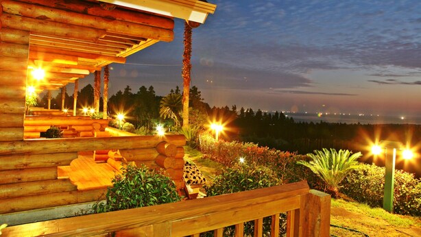 Uma cabana de madeira com varanda, rodeada por jardins bem cuidados e vista para a cidade ao entardecer.