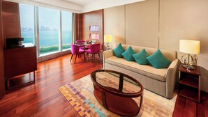 Quarto de hotel moderno com uma grande janela, um sofá com almofadas azul-turquesa, uma mesa de centro de vidro e um suporte de TV de madeira.