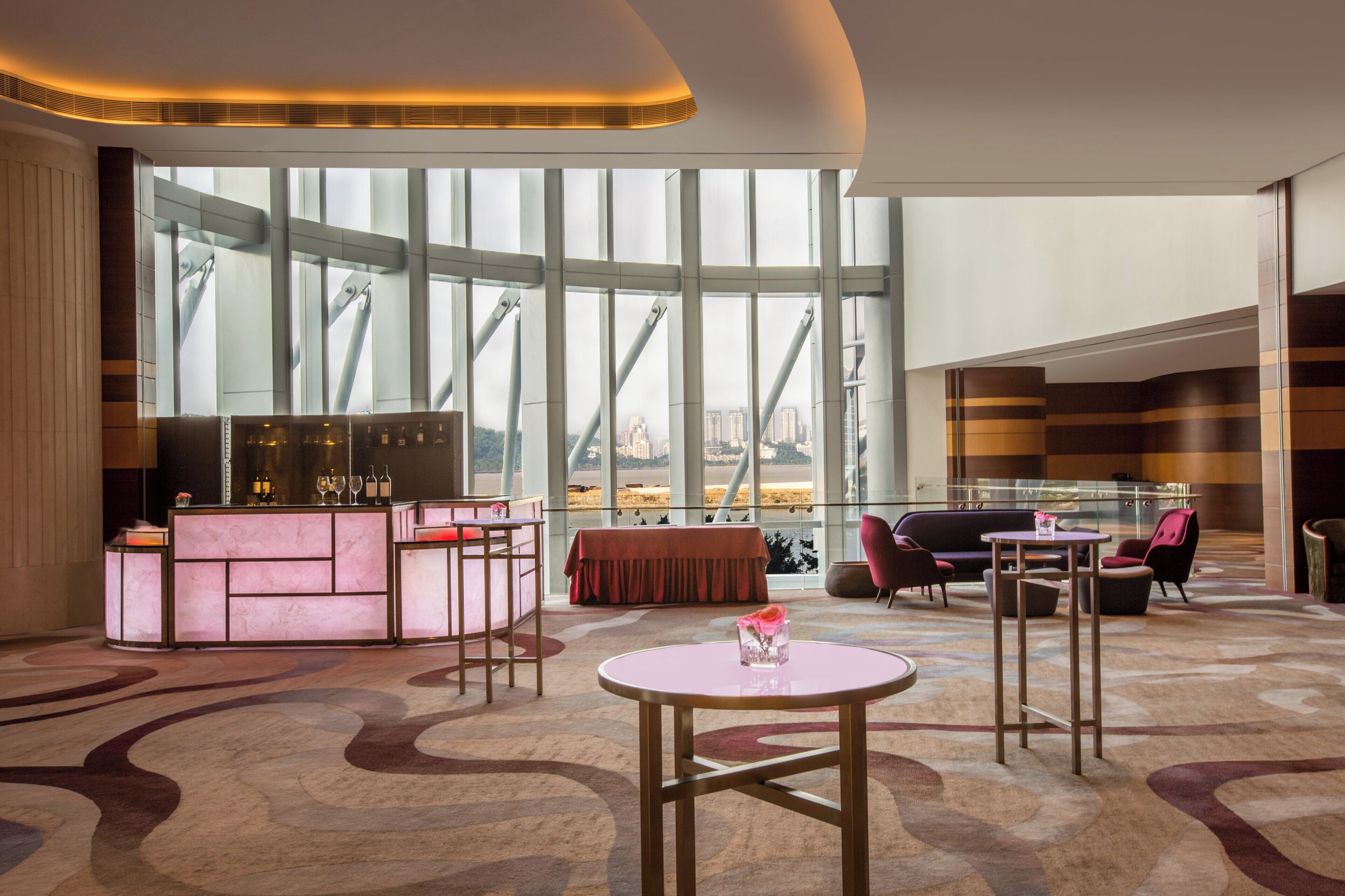 Um hall de hotel moderno com área de bar, assentos e grandes janelas que oferecem vista de uma área de construção.