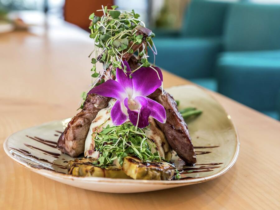 Prato de comida com carne grelhada, legumes e uma flor roxa para decorar.