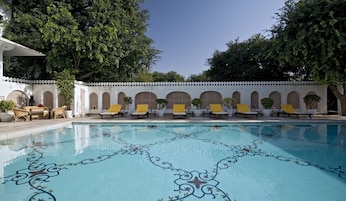 Uma piscina com design de azulejos geométricos, rodeada por paredes brancas com janelas em arco e espreguiçadeiras amarelas.