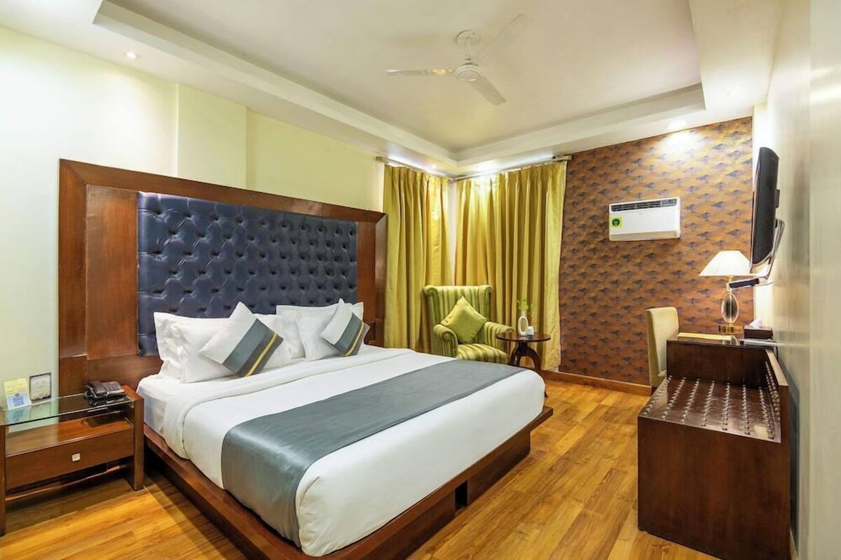 Quarto de hotel com uma cama grande, cabeceira de madeira, escrivaninha com luminária, cadeira e televisão.