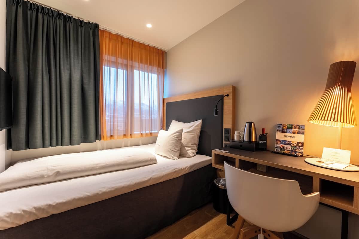 Quarto de hotel com uma cama, uma mesa com abajur, uma cadeira, uma televisão e uma janela com cortinas.