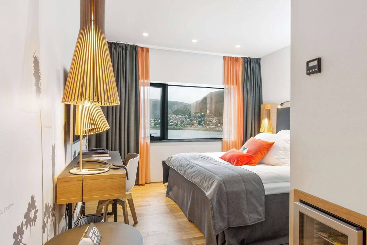 Quarto de hotel com cama, escrivaninha, cadeira, abajur e vista para a cidade pela janela.