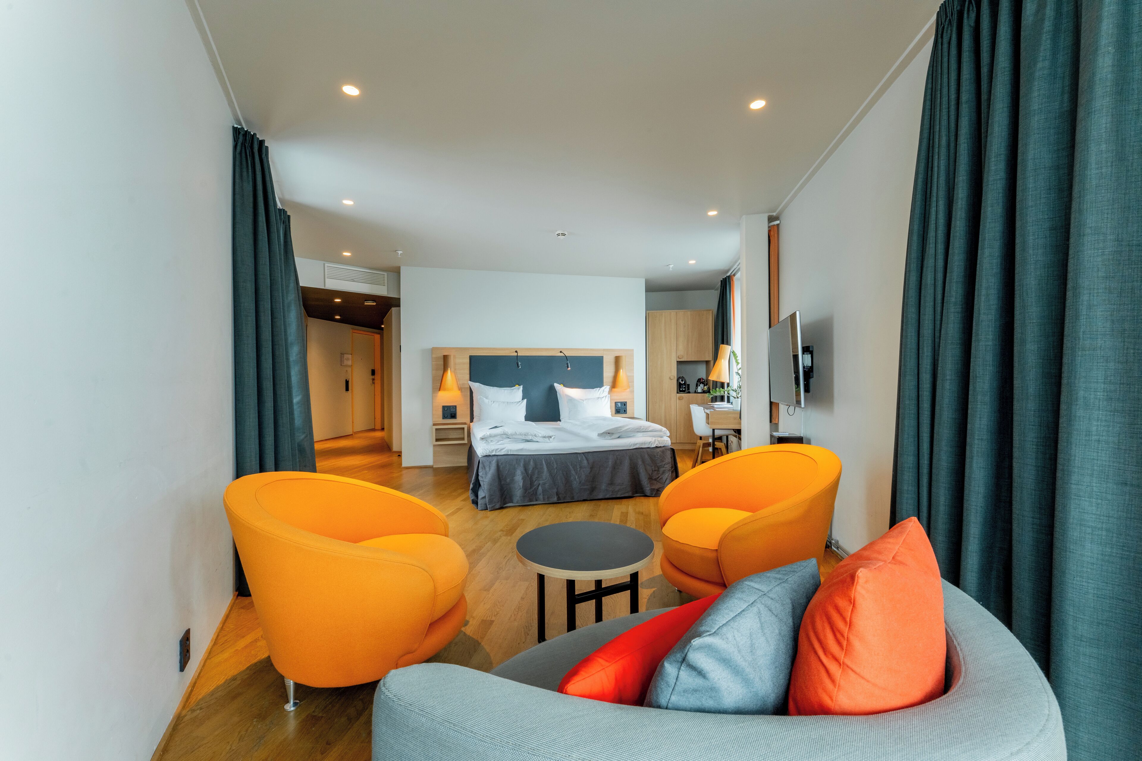 Quarto de hotel moderno com uma cama, duas poltronas laranja, um sofá cinza, uma mesa redonda pequena e uma televisão.