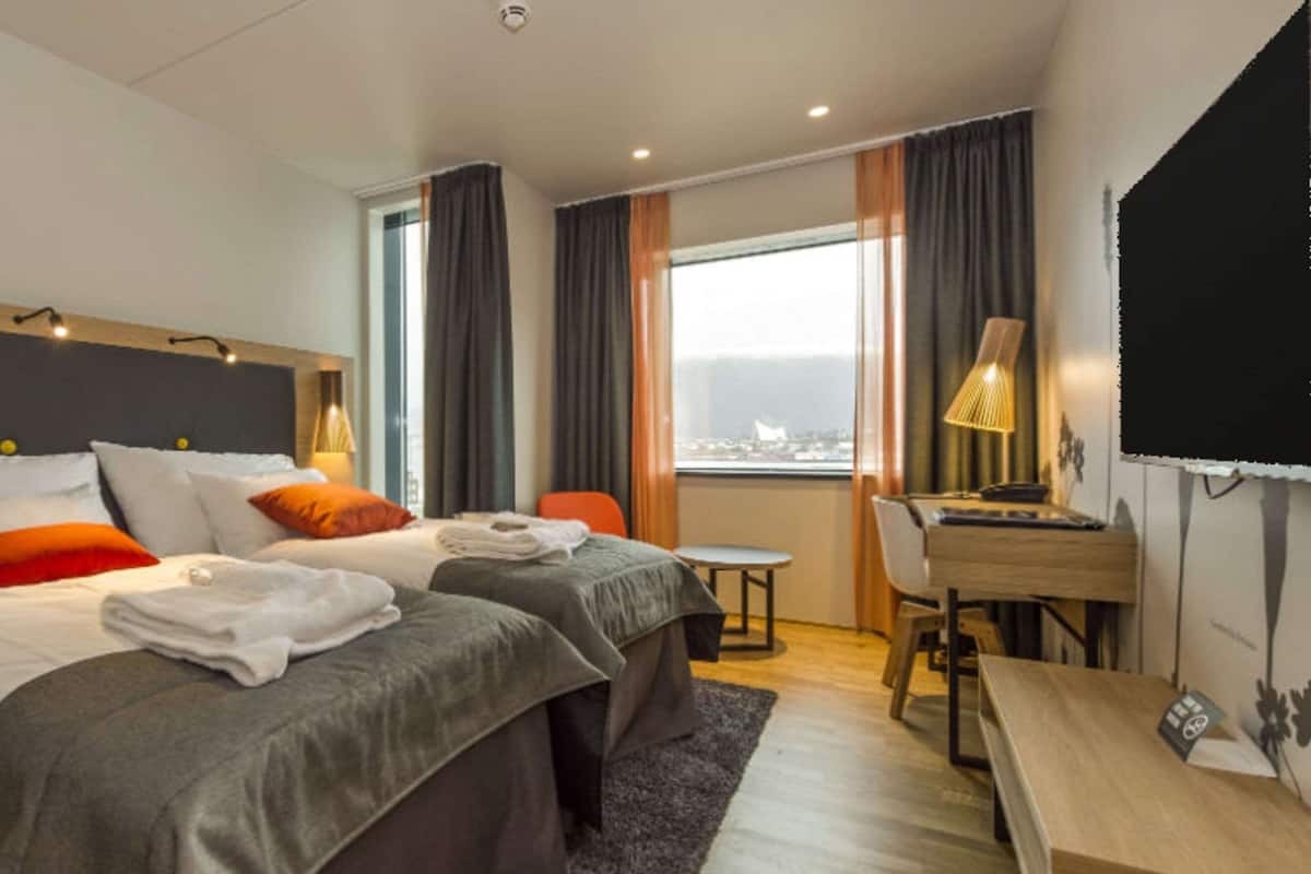 Quarto de hotel com duas camas, uma escrivaninha, uma cadeira, um abajur e uma janela grande com vista.