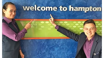 Uma placa de boas-vindas com a inscrição 'welcome to hampton' e duas pessoas apontando para ela.