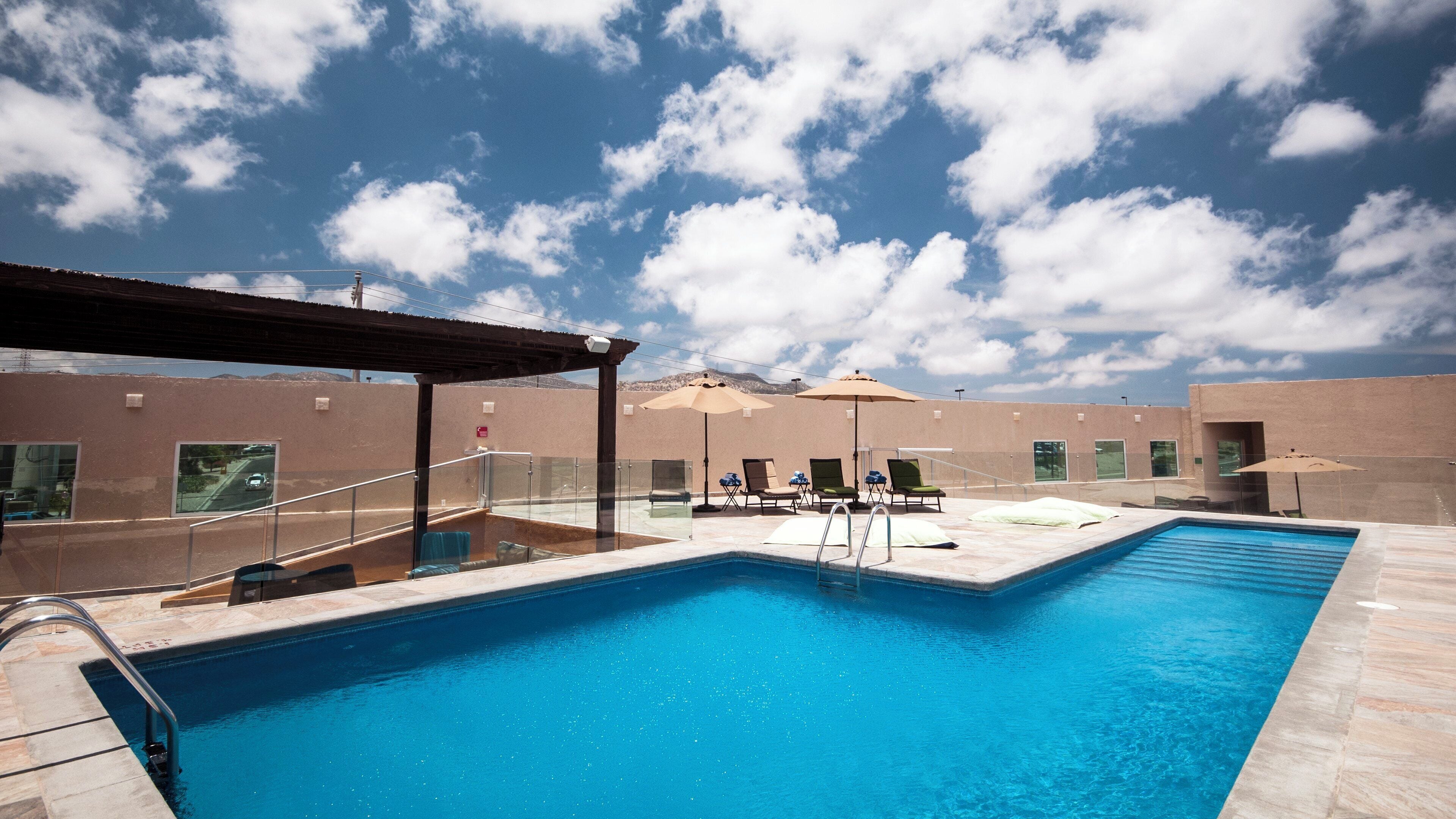 Piscina com espreguiçadeiras e guarda-sóis, cercada por um edifício com grandes janelas.