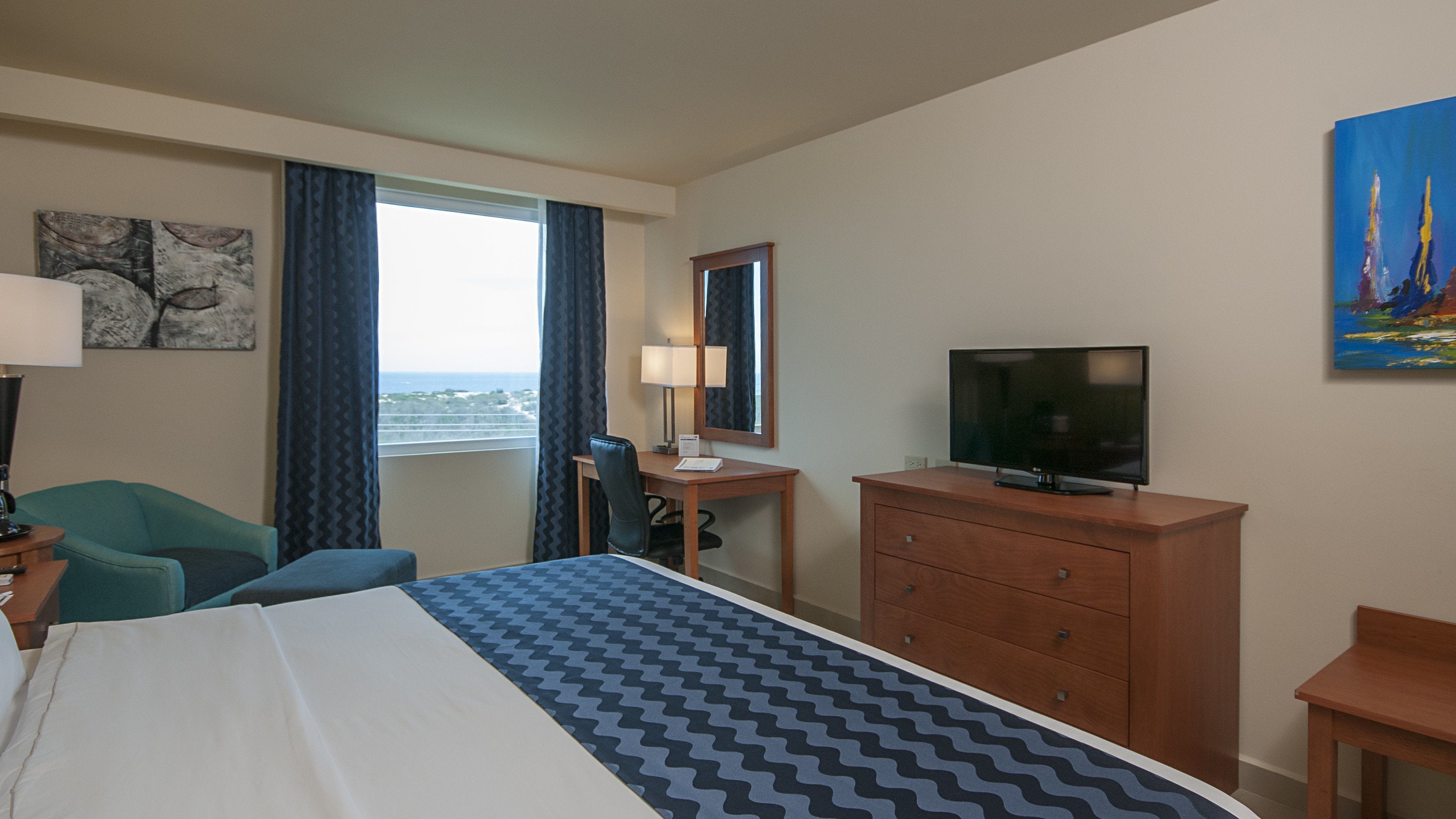 Quarto de hotel com cama, escrivaninha com cadeira, televisão, cómoda e obras de arte nas paredes.