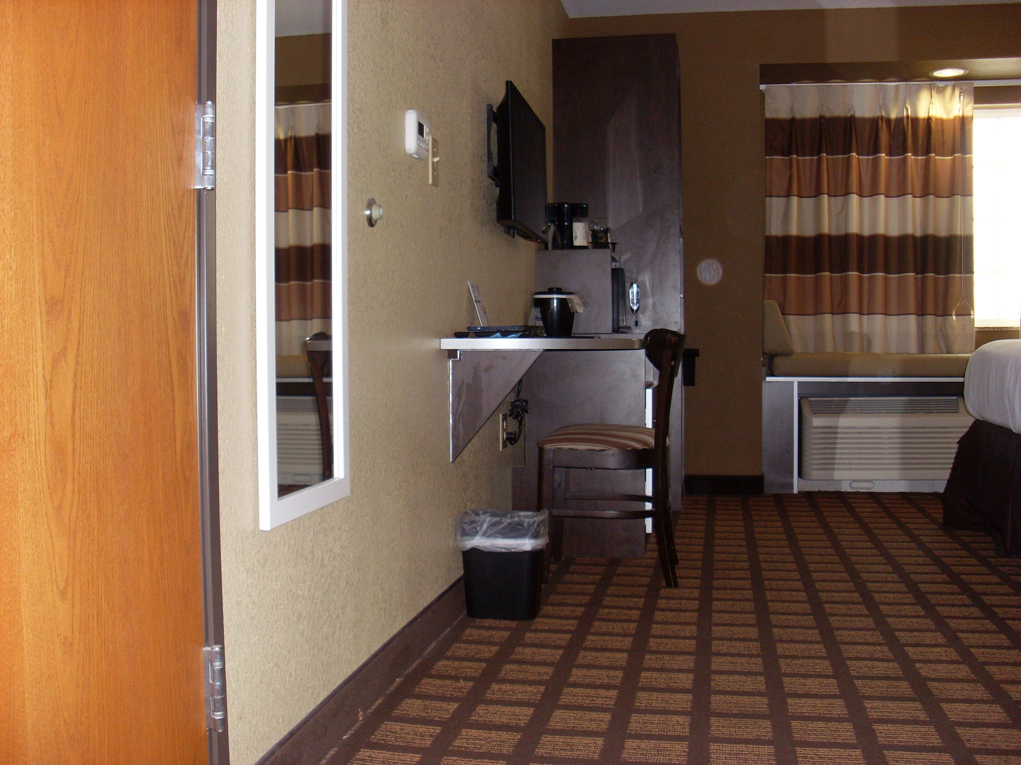 Quarto de hotel com porta de madeira, pequeno espaço de cozinha, uma cadeira, lixeira e tapete estampado.