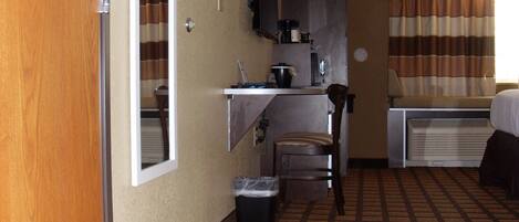 Quarto de hotel com porta de madeira, pequeno espaço de cozinha, uma cadeira, lixeira e tapete estampado.