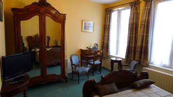 Quarto de hotel com um espelho grande, uma cama, uma escrivaninha com cadeira, televisão e uma janela com cortinas.