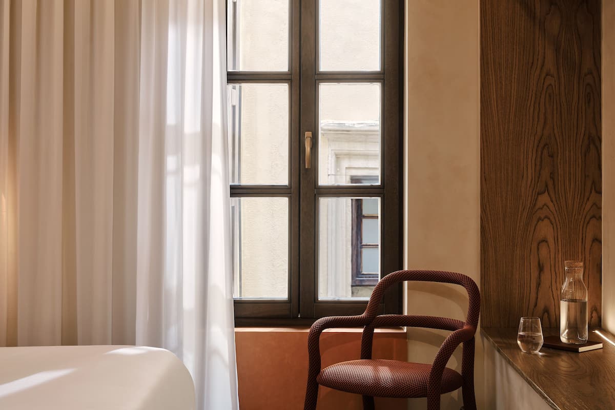 Quarto de hotel com uma janela grande, uma cadeira, uma mesa de cabeceira com garrafa e copo, e uma cama.