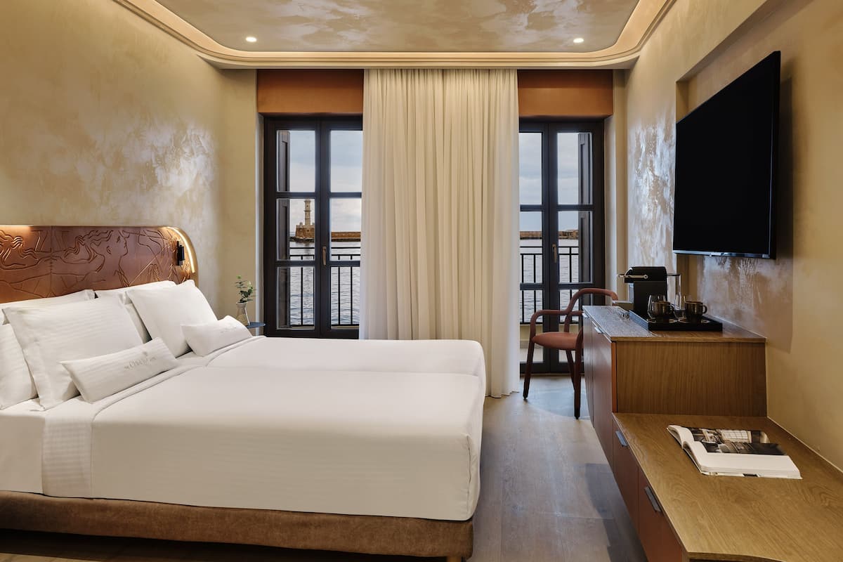 Quarto de hotel com uma cama grande, uma escrivaninha com cadeira, uma televisão e vista para a água através de uma janela.