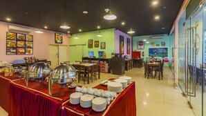 Disposição de buffet com diversas estações de serviço, um balcão com caixa registradora e uma área de estar com mesas e cadeiras.