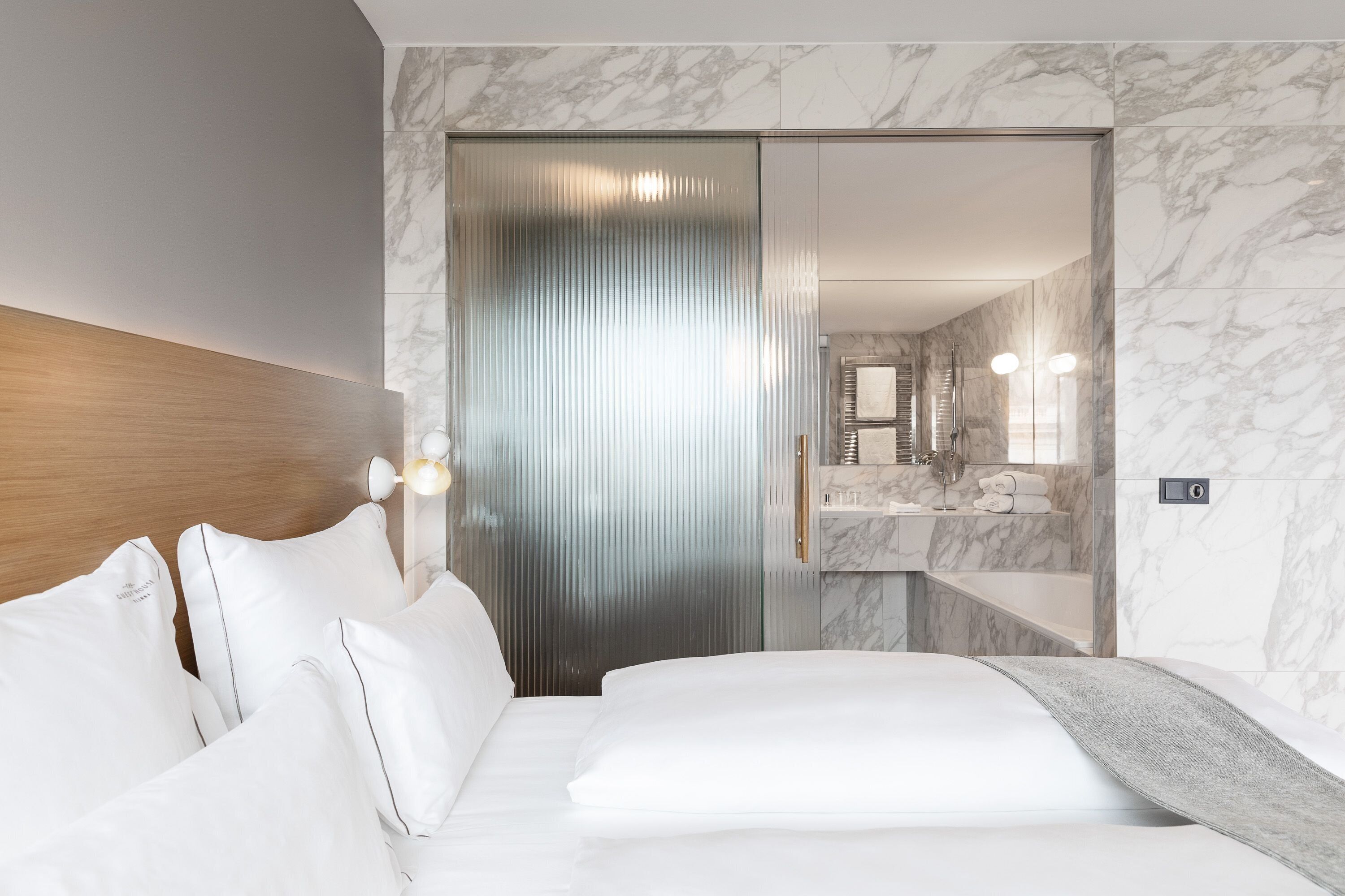Quarto de hotel moderno com uma cama grande, cabeceira de madeira, porta de vidro deslizante para um banheiro e paredes de mármore.