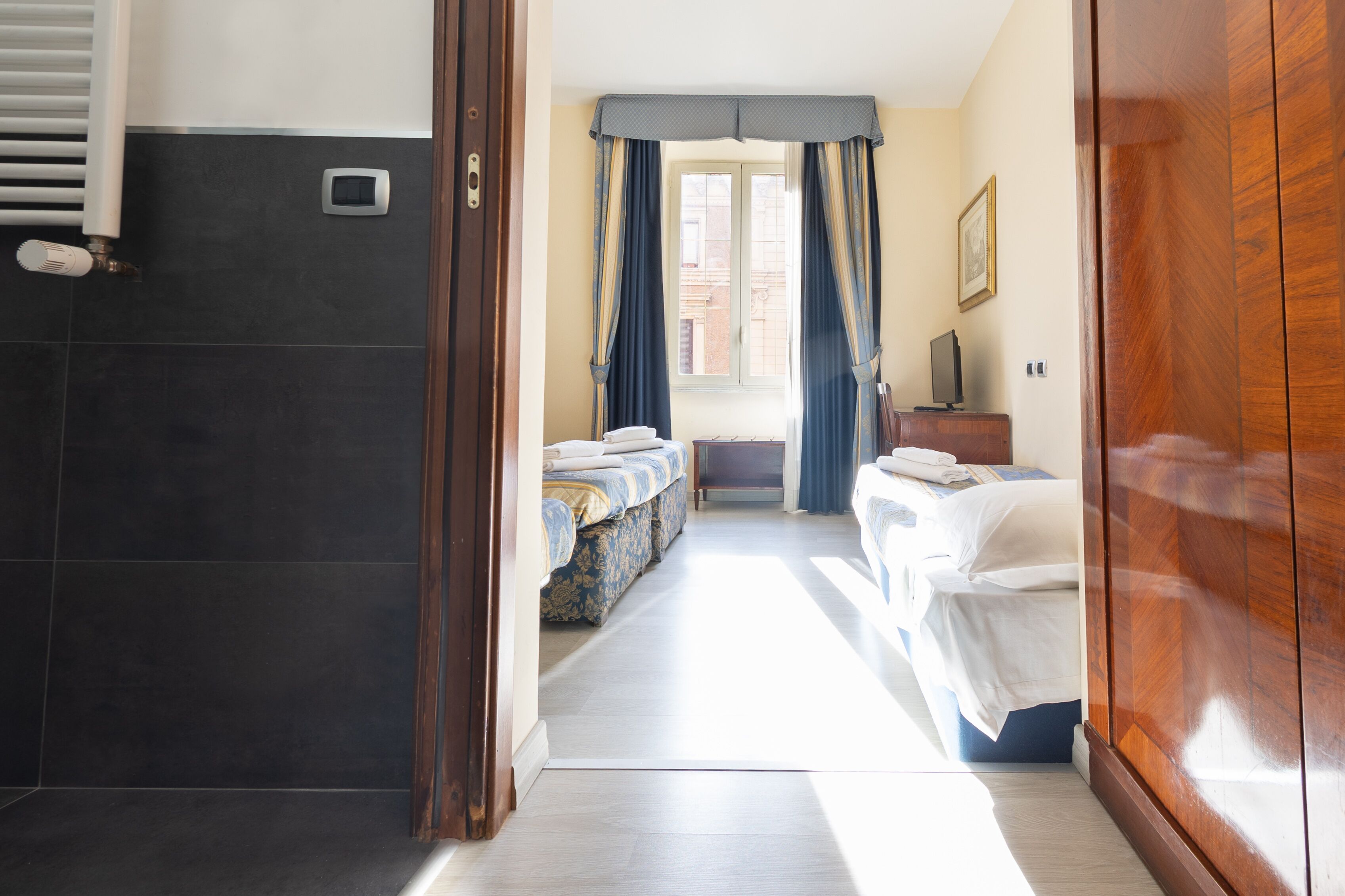 Quarto de hotel com cama, mesa de cabeceira, janela com cortinas e porta.