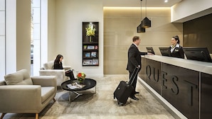 Um lobby moderno de hotel com um balcão de recepção identificado como “QUEST”, uma área de estar com uma mesa de centro e cadeiras, e uma estante de livros com livros e um vaso de flores.