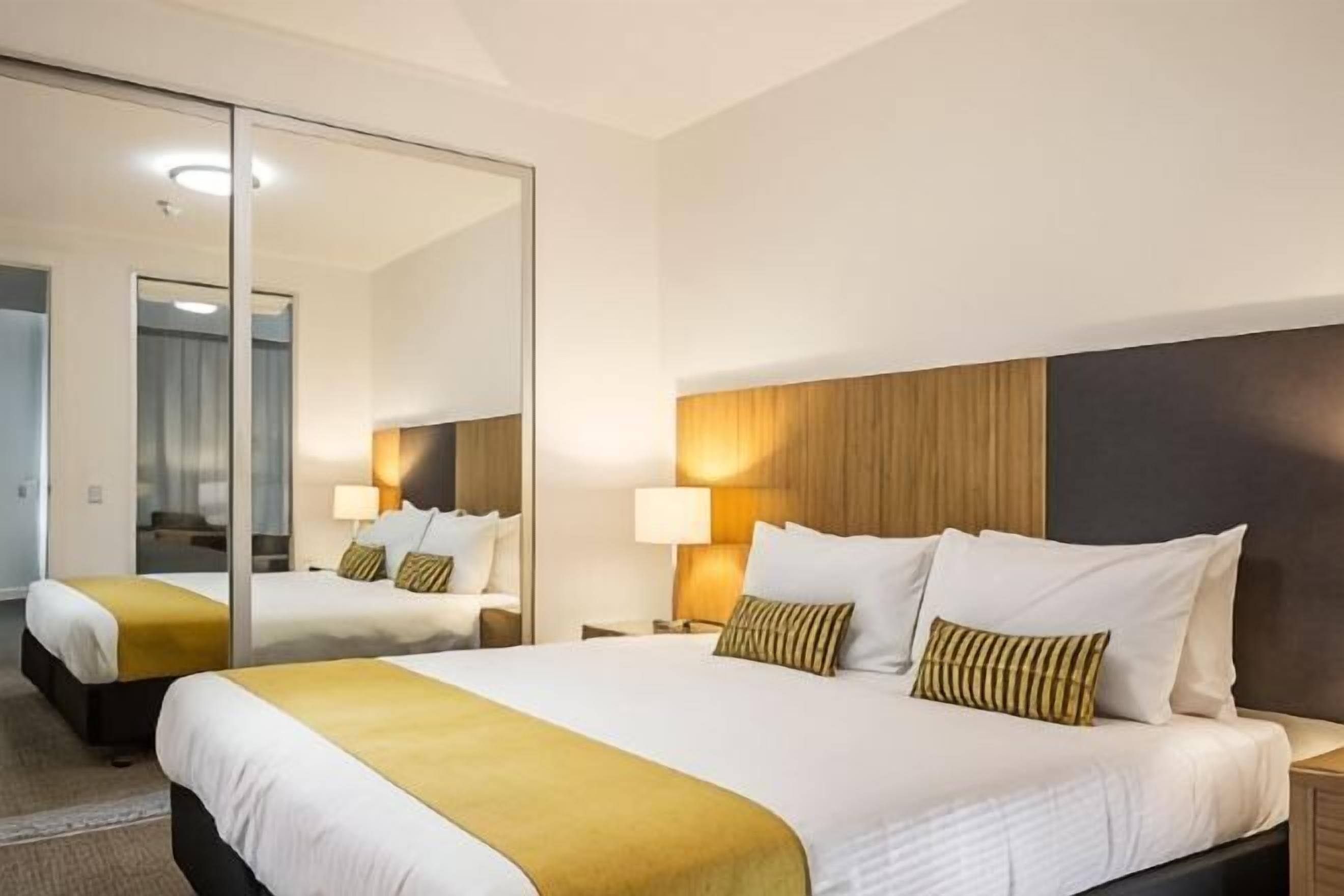 Quarto de hotel com duas camas, um espelho grande, abajures e cabeceira de madeira.