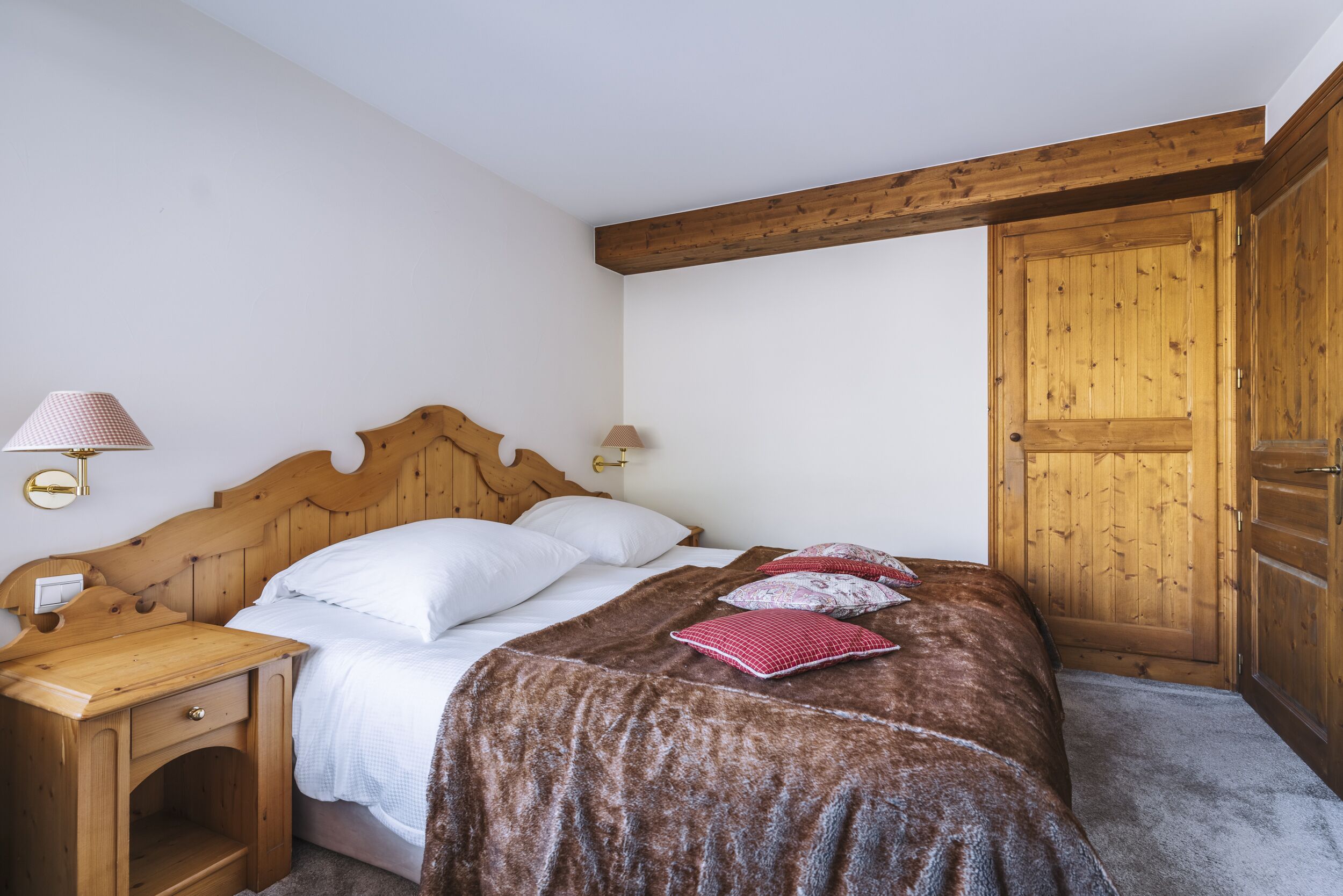 Quarto com cabeceira de madeira, guarda-roupa de madeira, mesa de cabeceira com luminária e cama com roupa de cama branca e manta marrom.