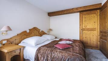 Quarto com cabeceira de madeira, guarda-roupa de madeira, mesa de cabeceira com luminária e cama com roupa de cama branca e manta marrom.