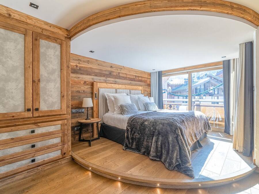 Quarto com teto de madeira arqueado, cama com manta escura, mesa de cabeceira com abajur e vista para um edifício tradicional lá fora.