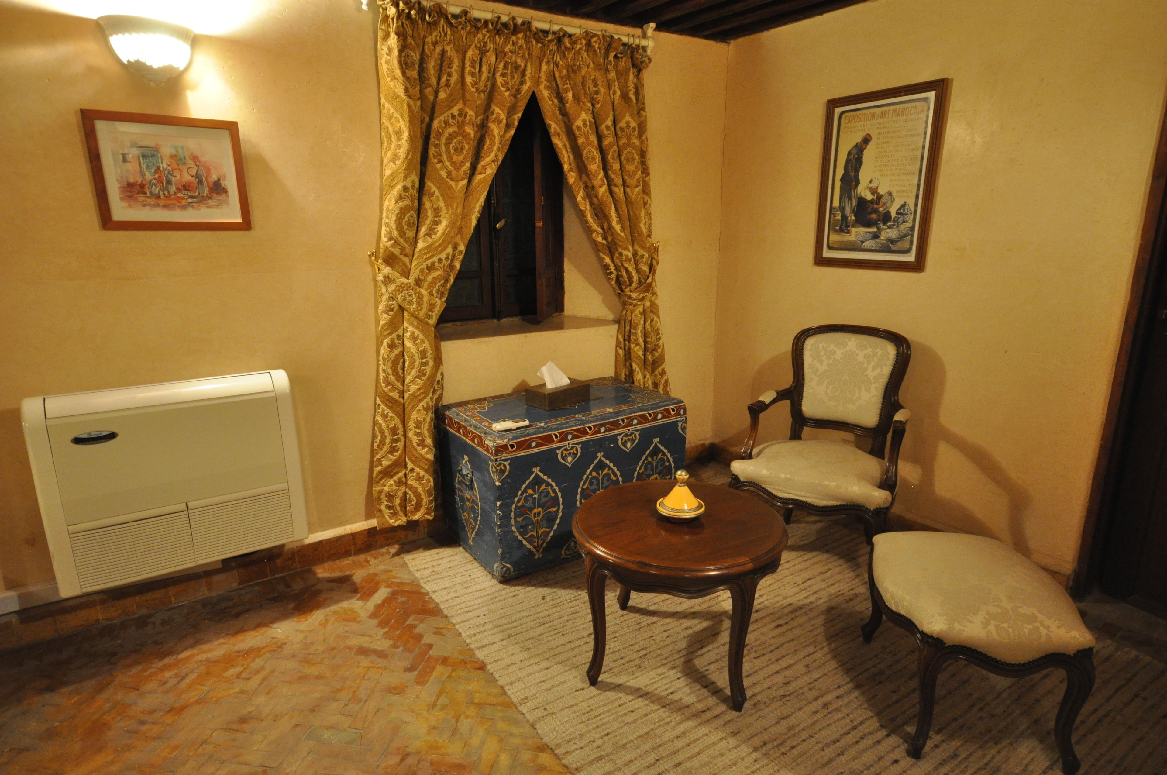 Um quarto com radiador, cortina estampada, um baú de madeira, mesa com luminária e uma cadeira.