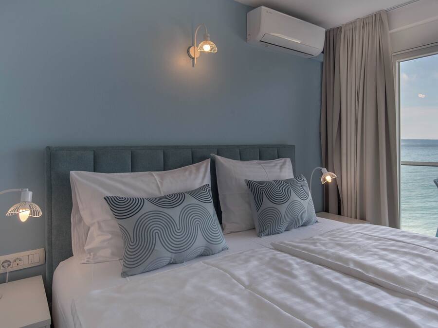 Uma cama bem arrumada com lençóis brancos e almofadas estampadas, uma mesa de cabeceira com abajur e vista para o mar através de uma grande janela.