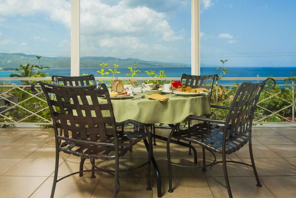Mesa posta para quatro pessoas, com cadeiras, toalha de mesa verde e vista para o oceano.