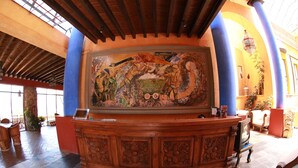 Um hall de entrada com um mural grande, balcão de recepção em madeira e piso de pedra.
