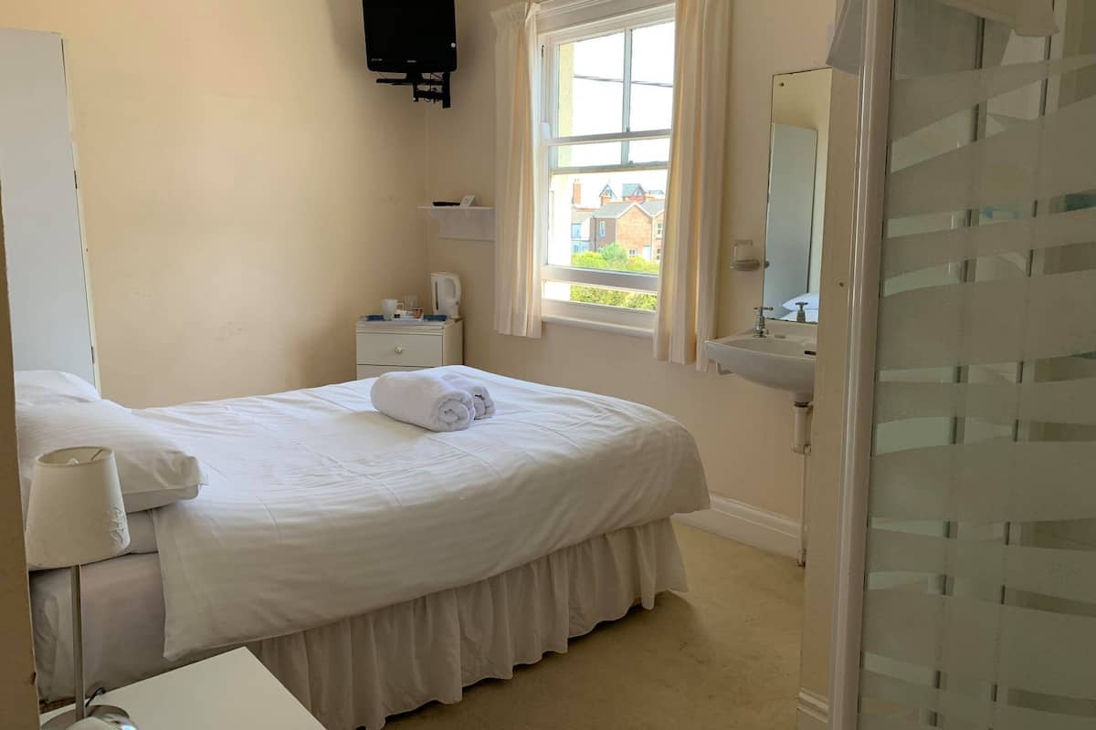 Quarto de hotel com cama, mesa de cabeceira com abajur, janela com cortinas, pia e espelho.