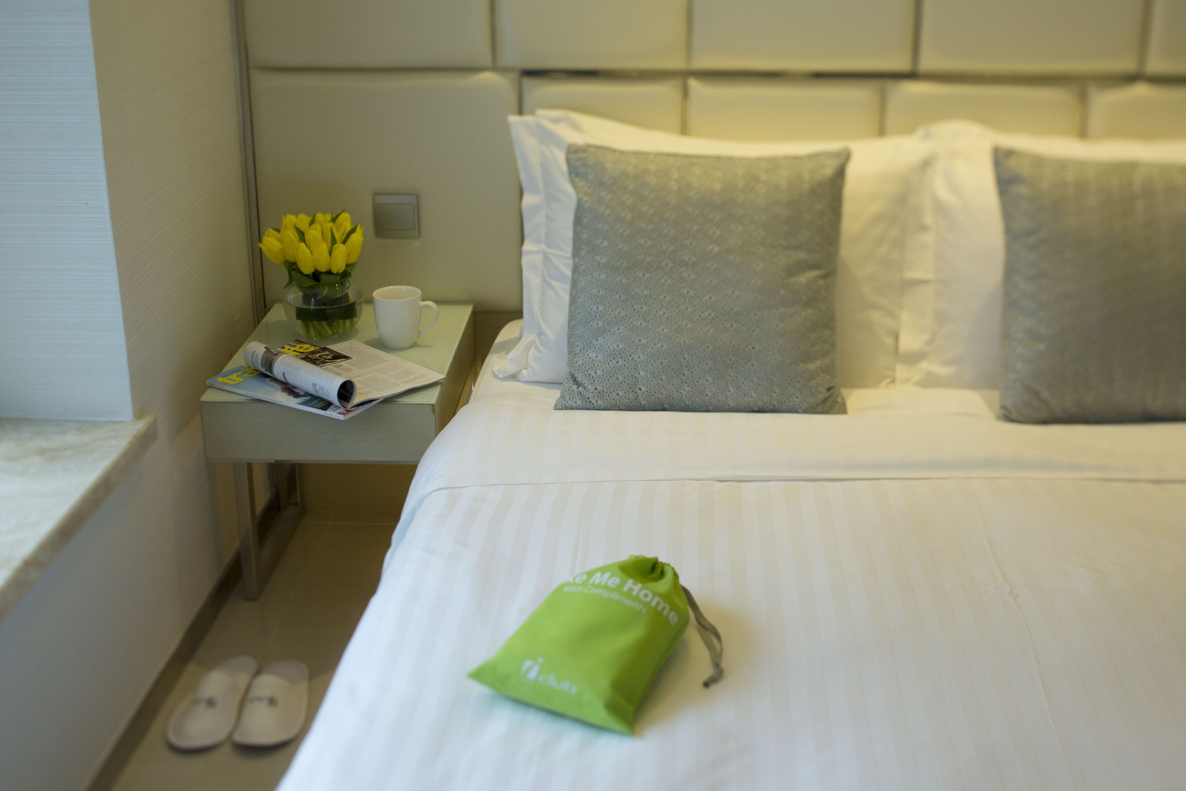 Quarto de hotel com cama bem arrumada, mesa de cabeceira com um vaso de flores amarelas, uma xícara e uma revista.