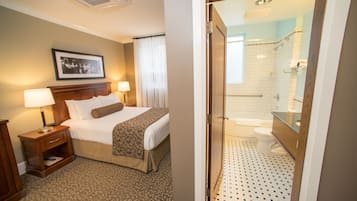 Quarto de hotel com cama bem arrumada, abajures, um quadro na parede e banheiro com piso ladrilhado e box de vidro.