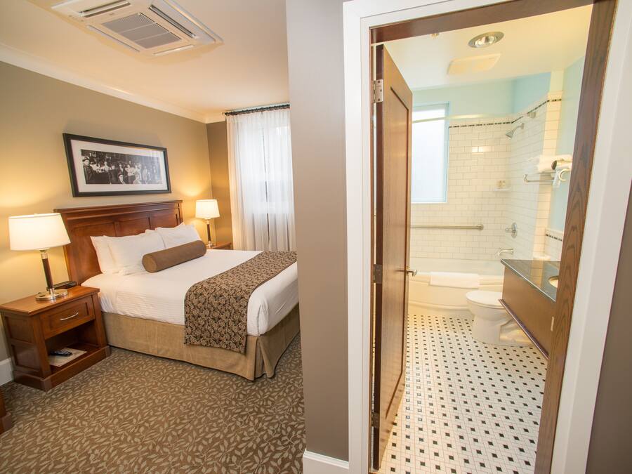 Quarto de hotel com cama bem arrumada, abajures, um quadro na parede e banheiro com piso ladrilhado e box de vidro.