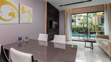 Uma sala de estar moderna com mesa de jantar de vidro, cadeiras brancas, sofá bege, televisão montada na parede e vista para uma piscina e áreas verdes lá fora.