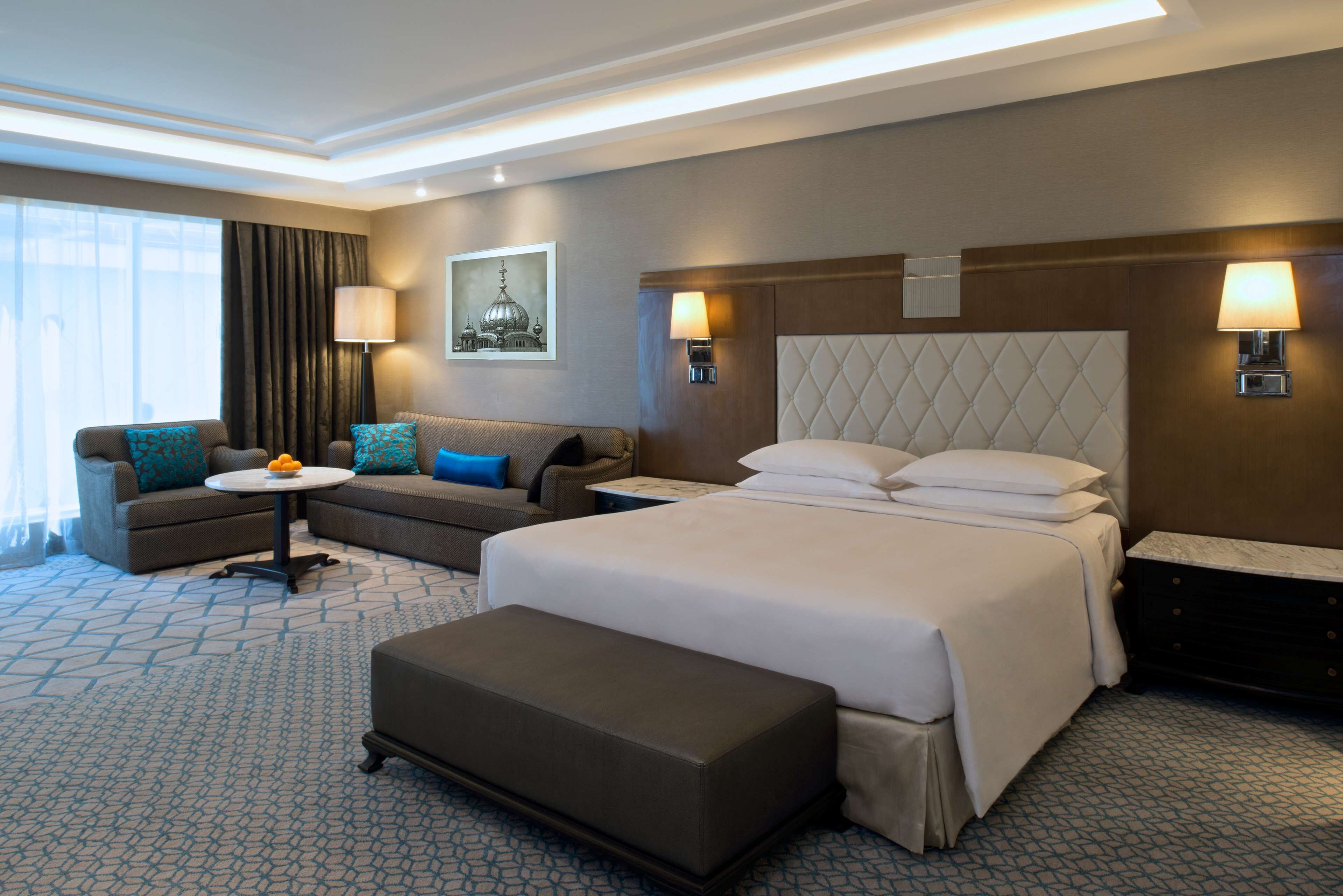 Quarto de hotel com uma cama grande, um sofá, uma mesinha e um banco. Há luminárias na parede e um quadro.