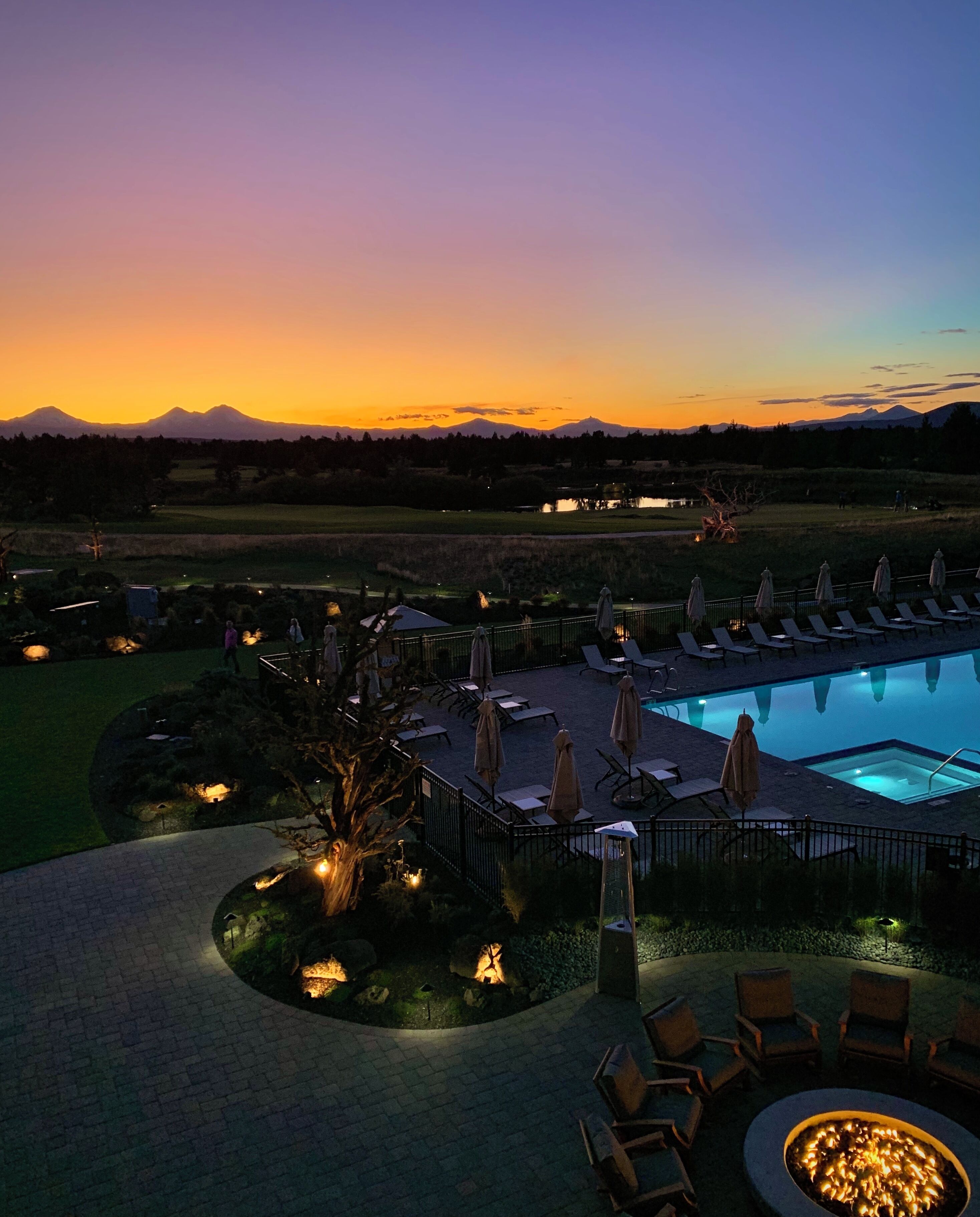 Uma área à beira da piscina com espreguiçadeiras e guarda-sóis, cercada por jardins bem cuidados e vista para montanhas ao longe durante o pôr do sol.