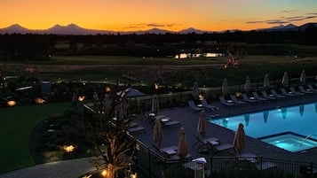 Uma área à beira da piscina com espreguiçadeiras e guarda-sóis, cercada por jardins bem cuidados e vista para montanhas ao longe durante o pôr do sol.