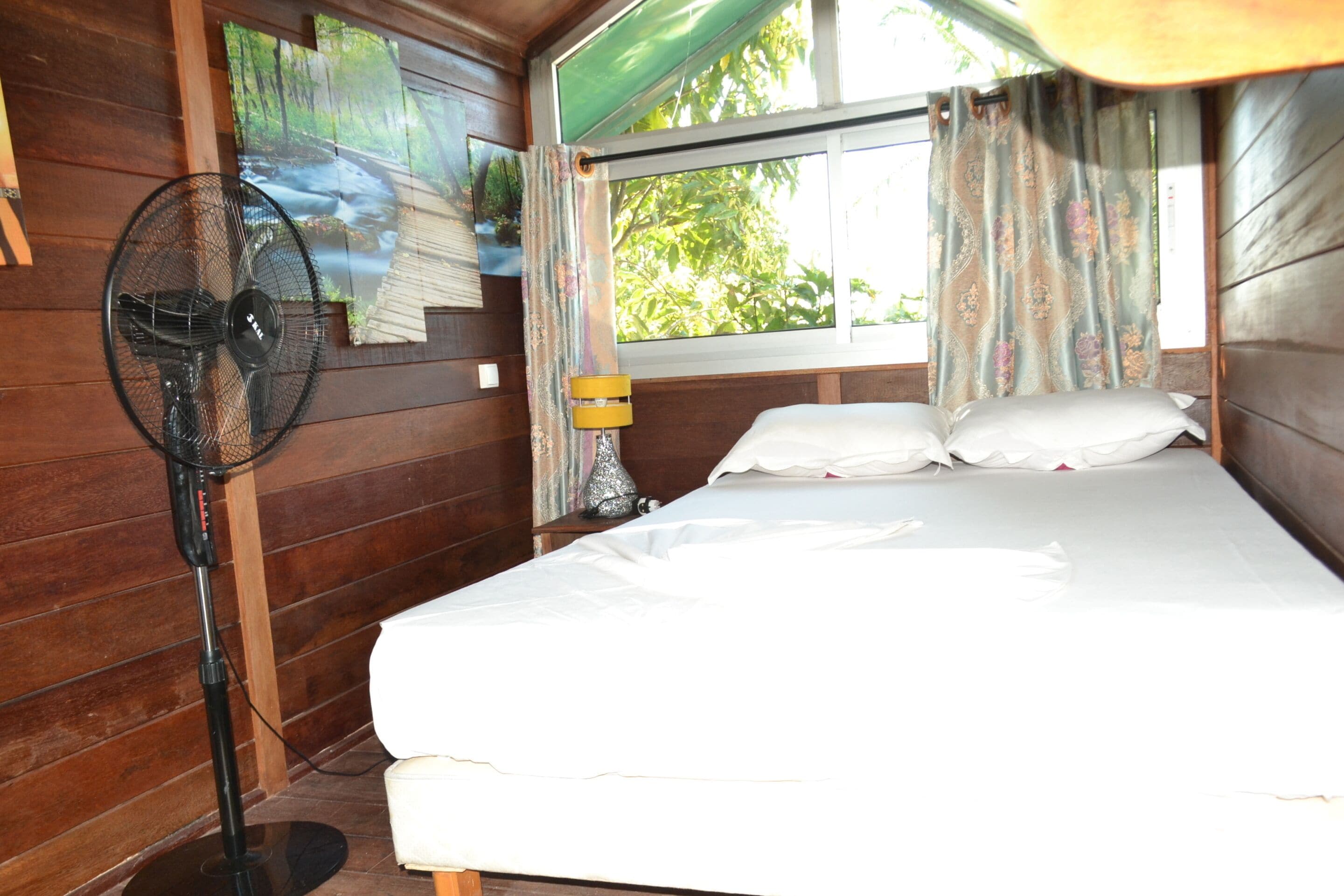 Um quarto com cama, ventilador, janela com cortinas e parede de madeira.