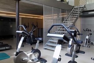 Uma academia com diversos equipamentos de exercícios, incluindo esteiras e elípticos, dispostos em um espaço amplo com escadas que levam a um andar superior.