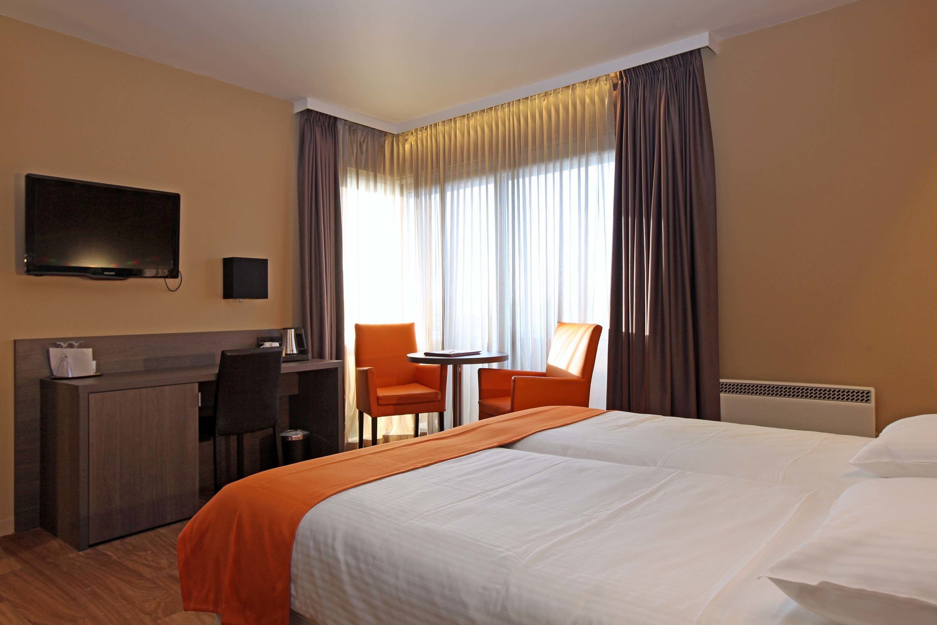 Quarto de hotel com cama, secretária com cadeira, televisão e janela com cortinas.