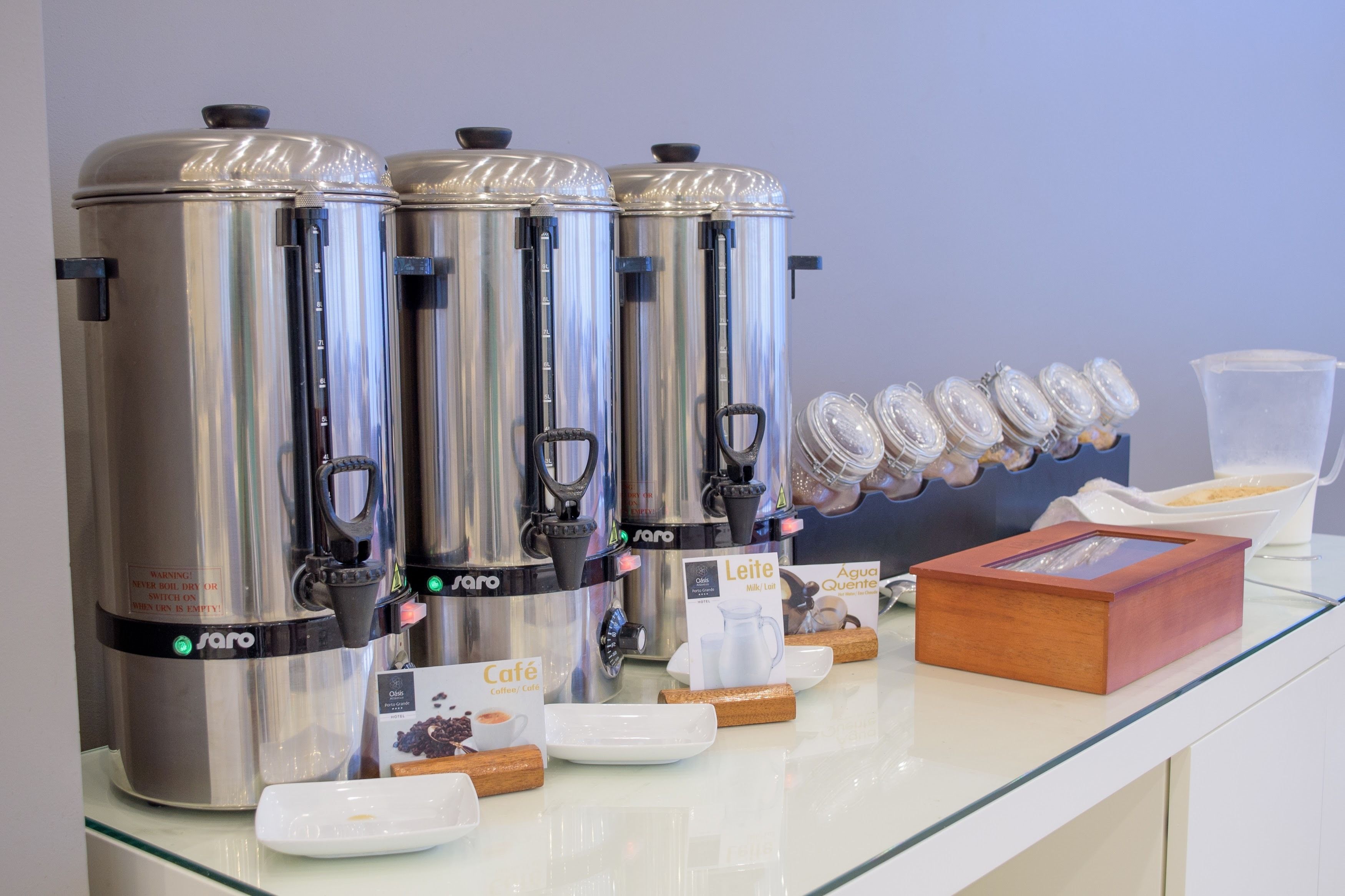 Um espaço para café com três dispensadores de café, identificados como 'Café' e 'Leite', e uma caixa de madeira com lenços de papel.